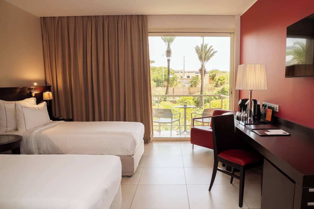 Quarto de hotel com duas camas, uma mesa com cadeira, TV e varanda com vista para palmeiras e edifícios.