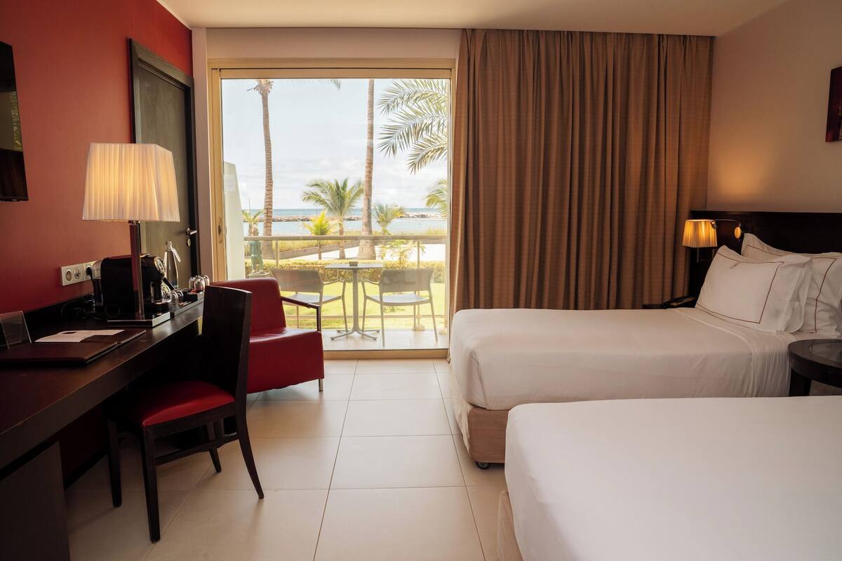 Quarto de hotel com duas camas, uma escrivaninha com cadeira, luminária e vista para a praia e palmeiras.