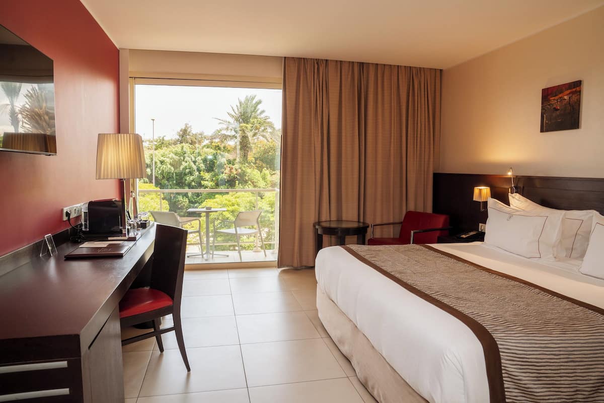 Quarto de hotel com cama, escrivaninha, cadeira e varanda com vista para o verde.