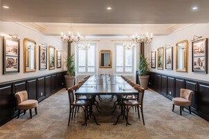 Uma sala de conferências com uma mesa comprida, cadeiras, lustres, quadros emoldurados e plantas em vasos.