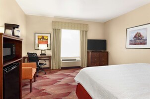 Quarto de hotel com cama, escrivaninha com cadeira, televisão, micro-ondas e frigorífico.