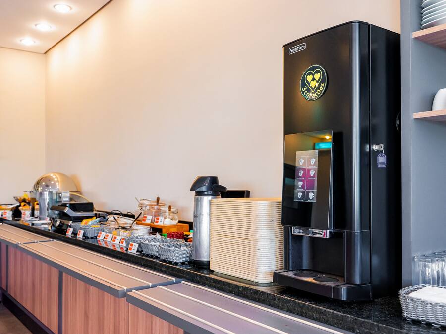 Uma estação de café com serviço próprio, com uma máquina de café, pilha de copos descartáveis e diversos acessórios para preparar café.