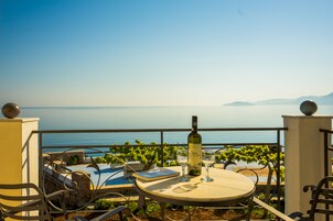 Um terraço com mesa, cadeiras, uma garrafa de vinho e um livro, com vista para um mar tranquilo e vegetação.