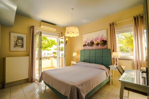 Quarto de hotel com cama, escrivaninha, cadeira, abajur, janela com cortinas, radiador e porta para área externa.