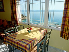 Uma área de refeições com uma mesa posta para dois, uma janela com vista para o mar e uma cadeira com capa xadrez.