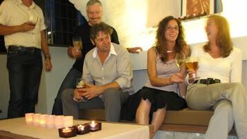 Um grupo de pessoas em uma sala com uma mesa posta com velas, algumas sentadas e outras em pé, indicando um encontro social.