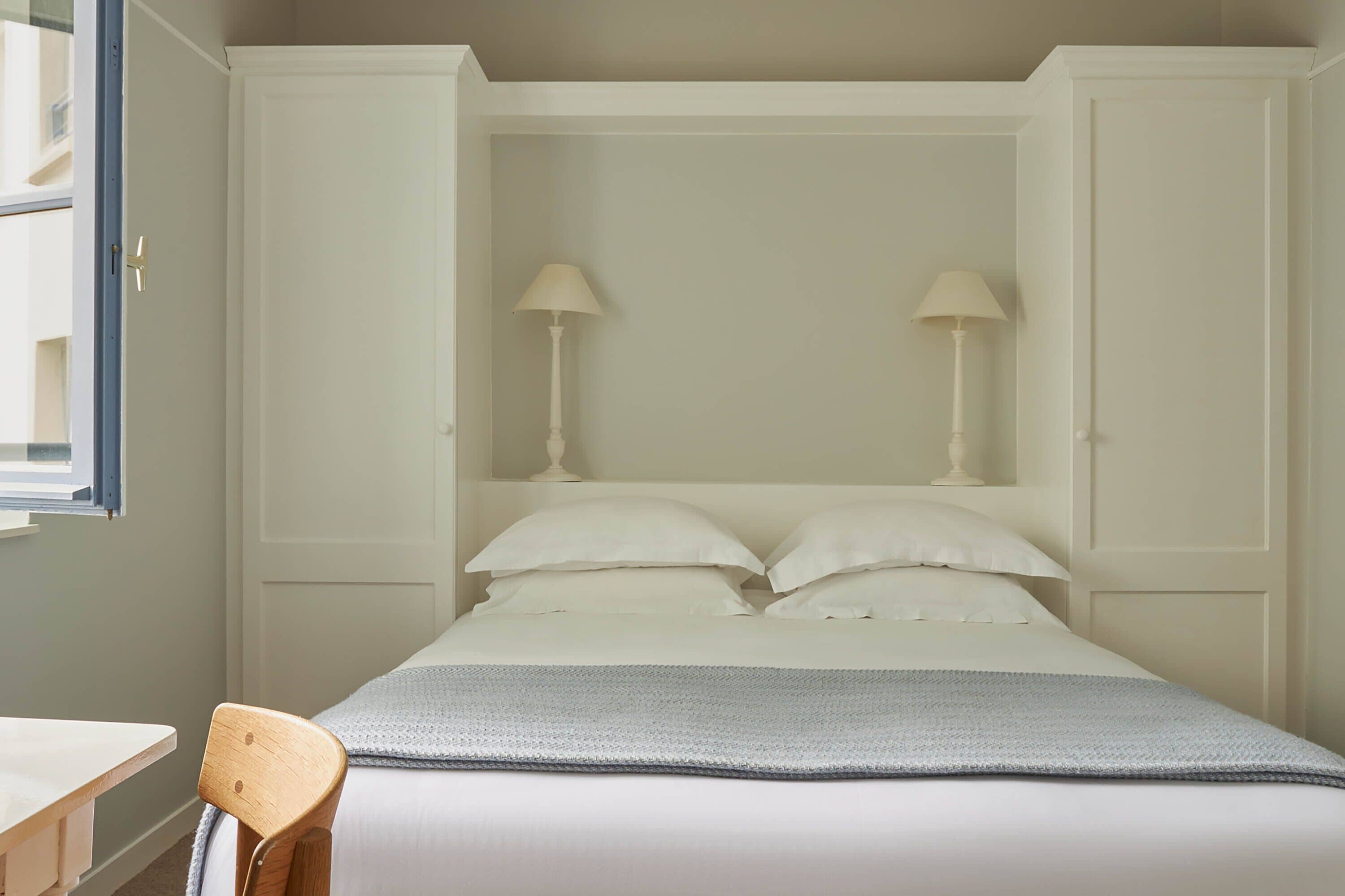 Quarto com cama, duas luminárias de cabeceira, uma cadeira e uma janela.