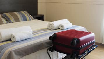 Uma cama com roupa de cama listrada e toalhas brancas dobradas, uma mala vermelha em um carrinho de bagagem, e uma mesa de cabeceira com uma luminária.