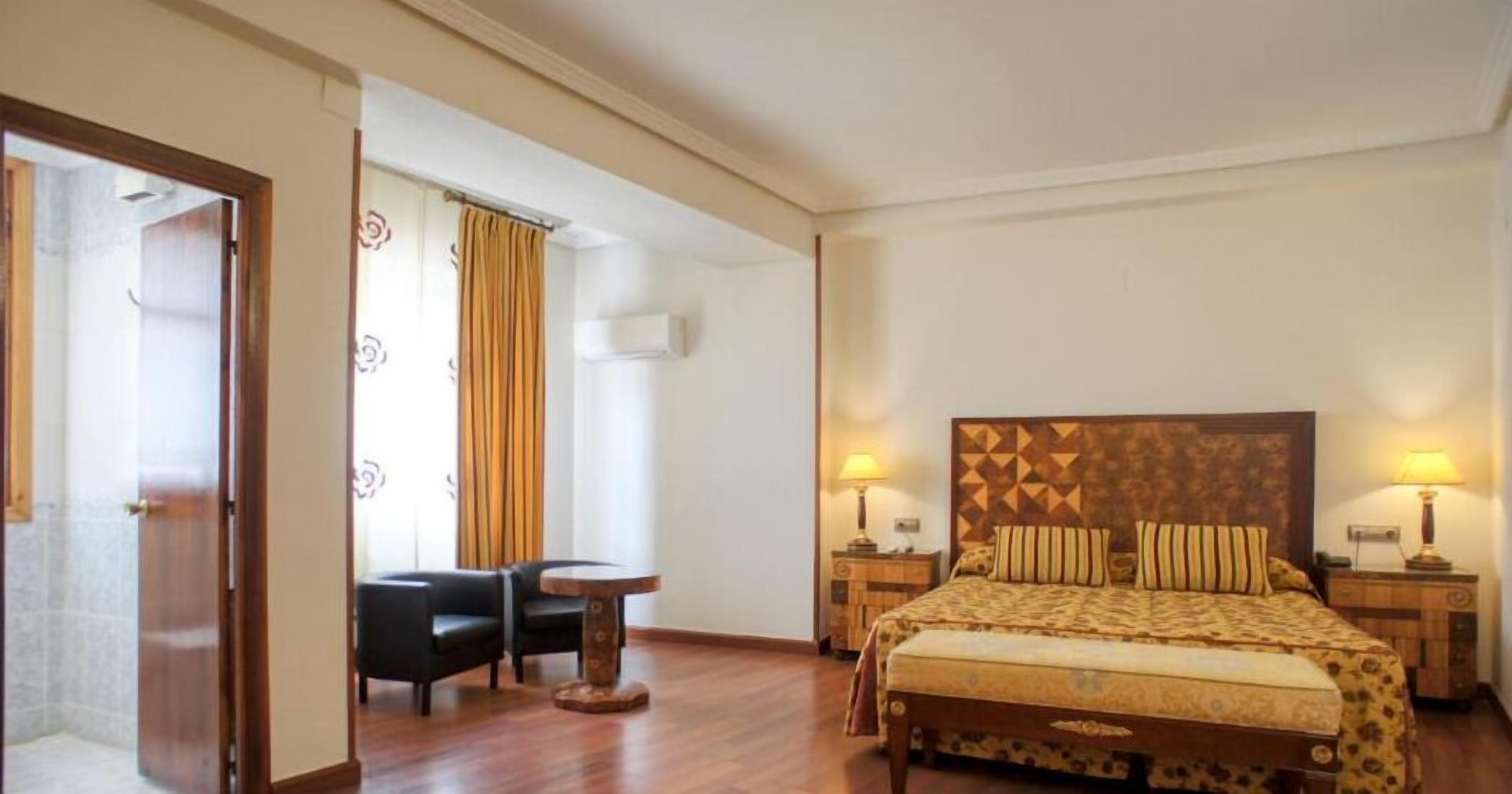 Quarto de hotel com uma cama grande, duas mesas de cabeceira com abajures, uma mesa redonda pequena com duas cadeiras e uma porta que dá para outro quarto.