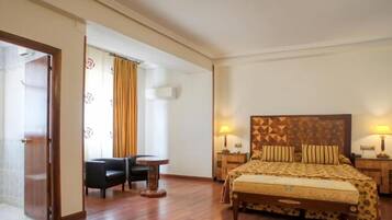 Quarto de hotel com uma cama grande, duas mesas de cabeceira com abajures, uma mesa redonda pequena com duas cadeiras e uma porta que dá para outro quarto.