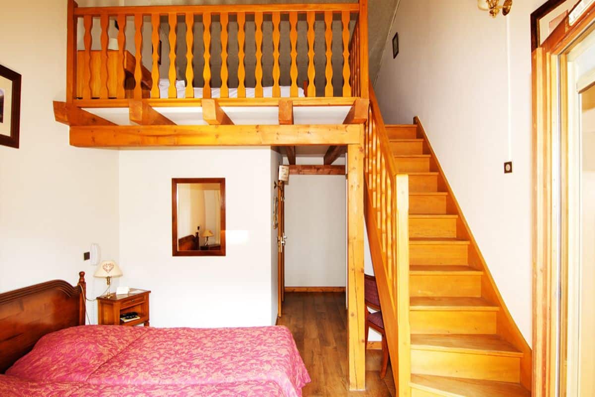 Quarto com cama de madeira, colcha rosa, escada de madeira e uma pequena mesa de cabeceira com abajur.