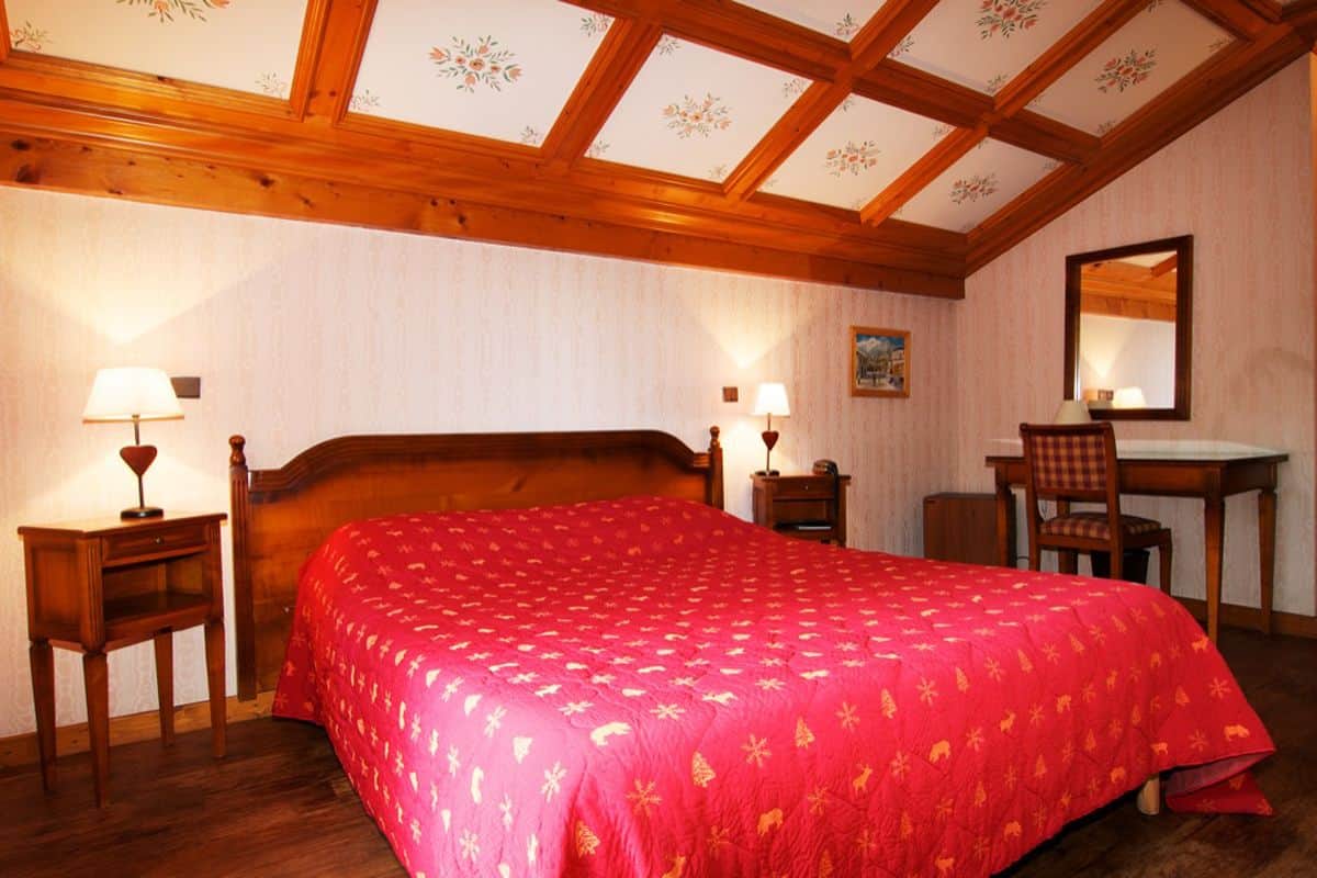 Quarto com teto de madeira, colcha vermelha, duas mesas de cabeceira com luminárias, uma cadeira e uma escrivaninha.