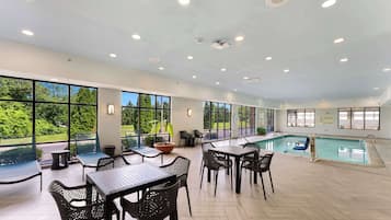 Área à beira da piscina com mesas e cadeiras, uma piscina e grandes janelas com vista para o verde.