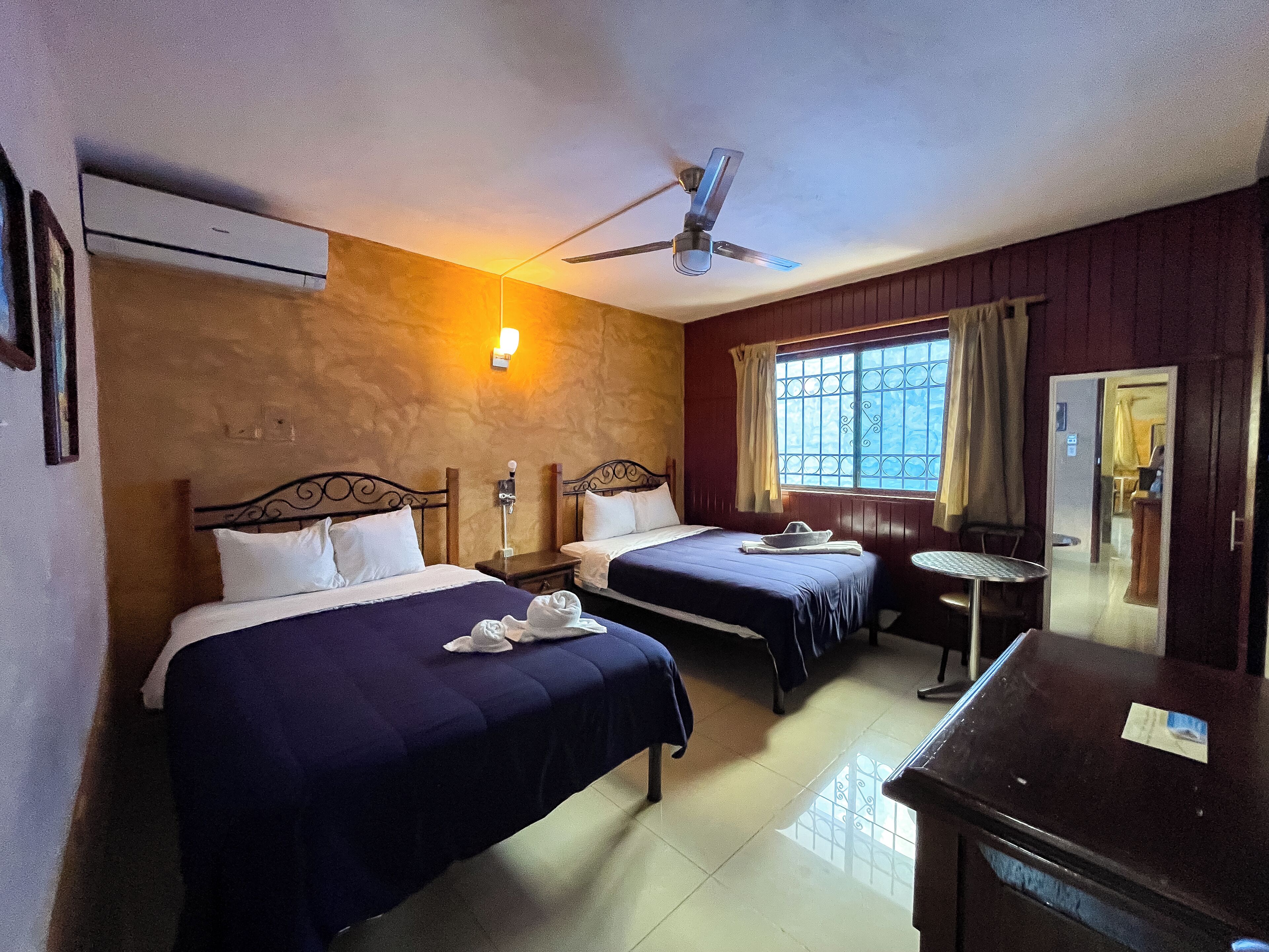 Quarto de hotel com duas camas, um ventilador de teto, uma janela com cortinas, uma pequena mesa e um espelho.