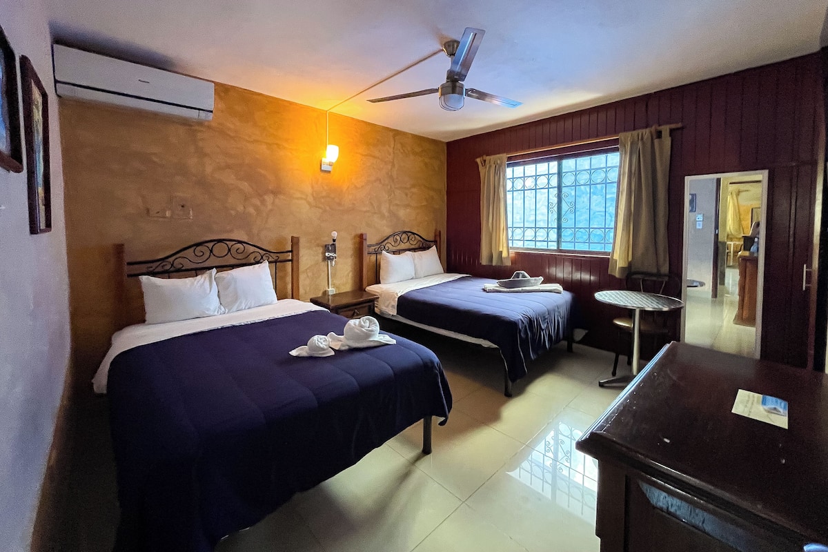 Quarto de hotel com duas camas, um ventilador de teto, uma janela com cortinas, uma pequena mesa e um espelho.