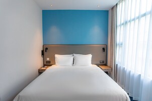 Quarto de hotel com uma cama grande, mesa de cabeceira e janela com cortinas.