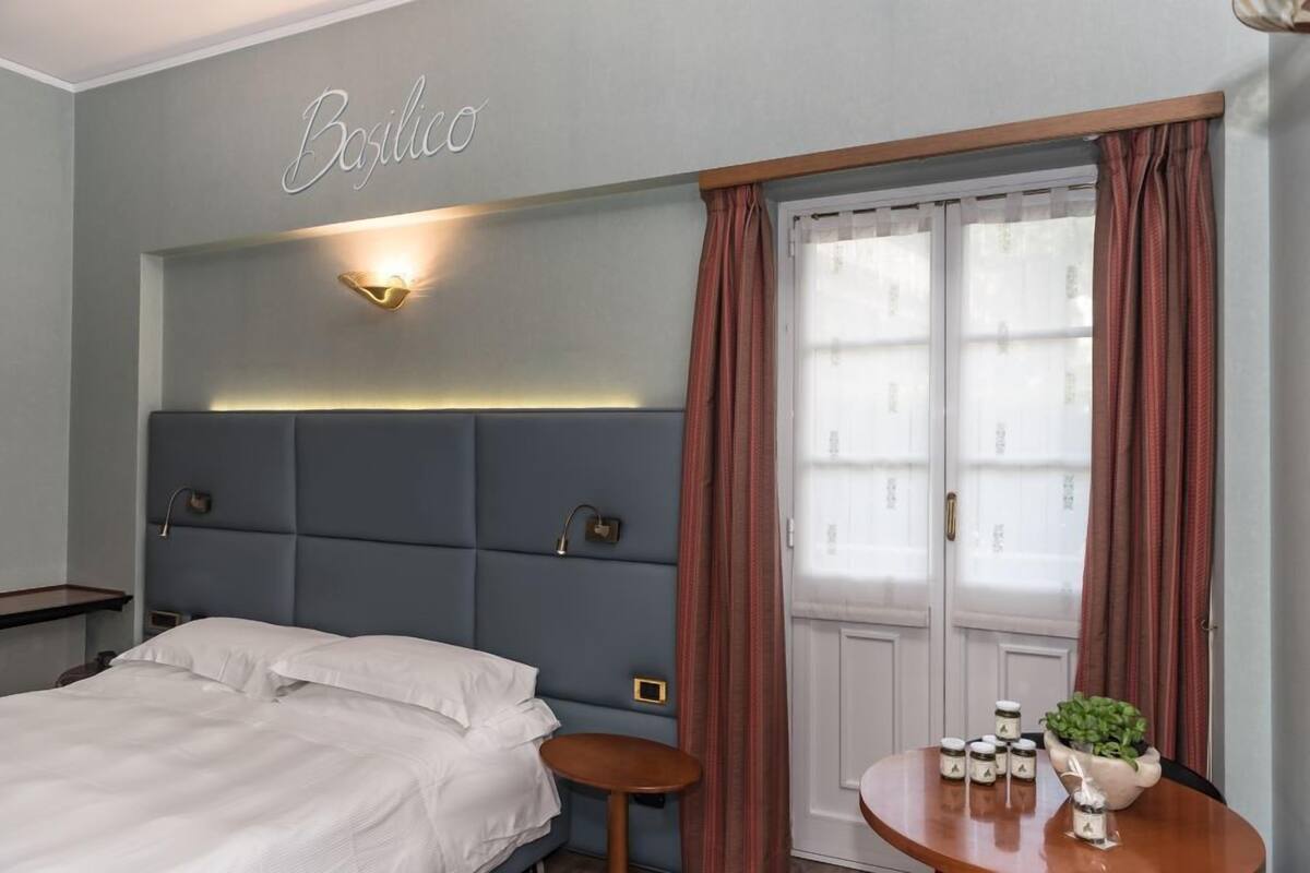 Quarto de hotel com cama, mesa de cabeceira, uma mesinha redonda com planta e velas, janela com cortinas e luminária na parede.