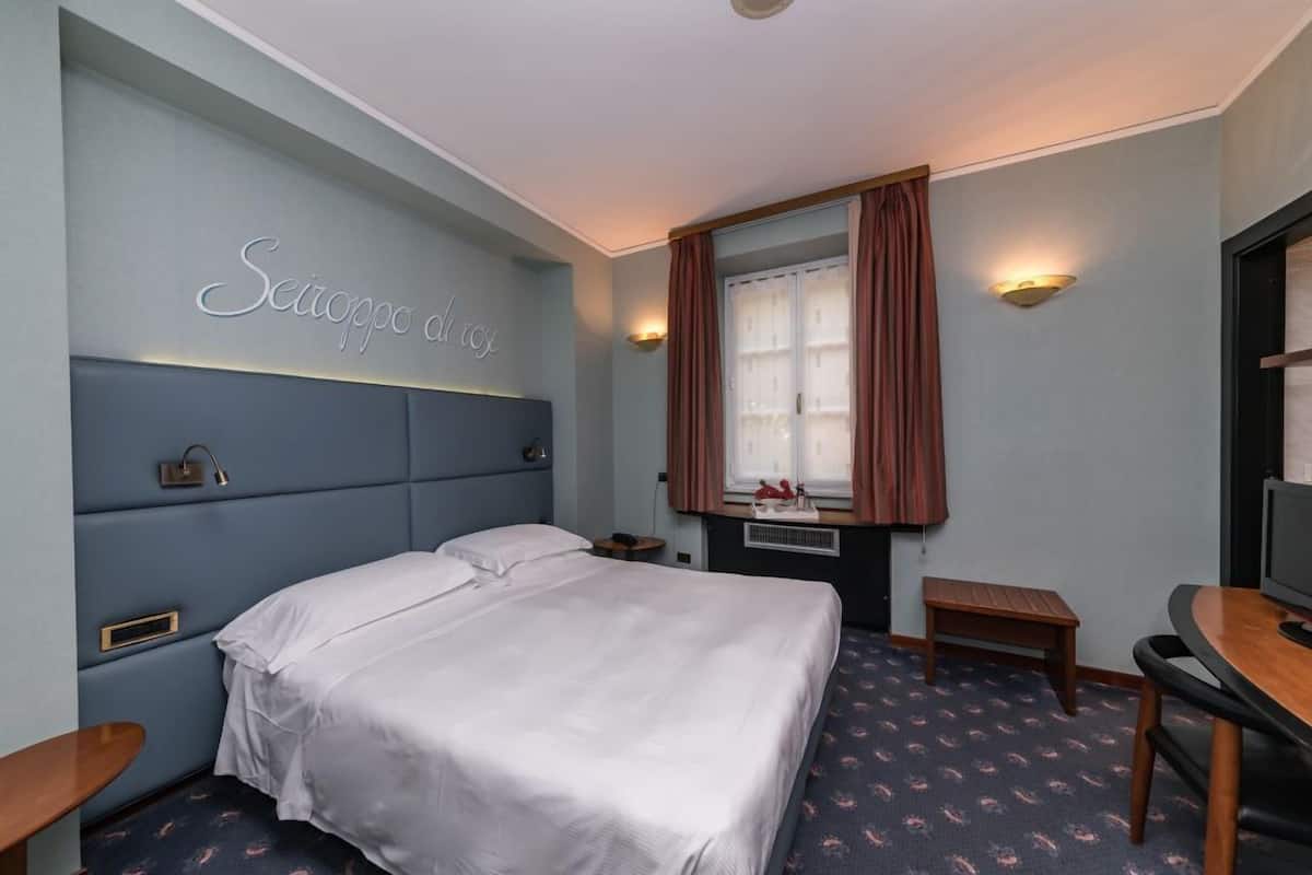 Quarto de hotel com cama, mesas de cabeceira, escrivaninha, cadeira, janela com cortinas, luminária na parede e placa sobre a cama.