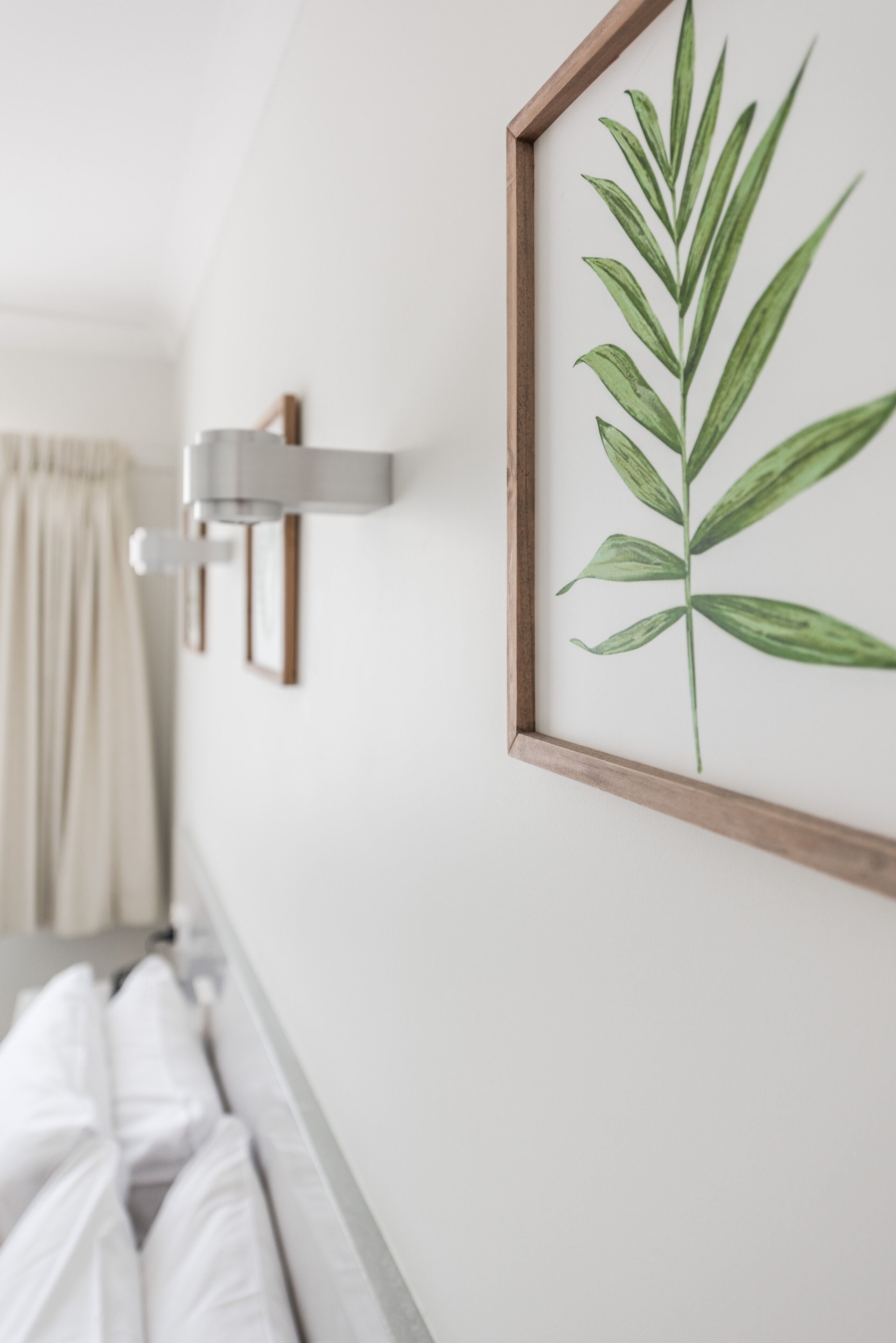 Uma impressão botânica emoldurada na parede, uma cama com roupa de cama branca e um candeeiro fixado na parede.