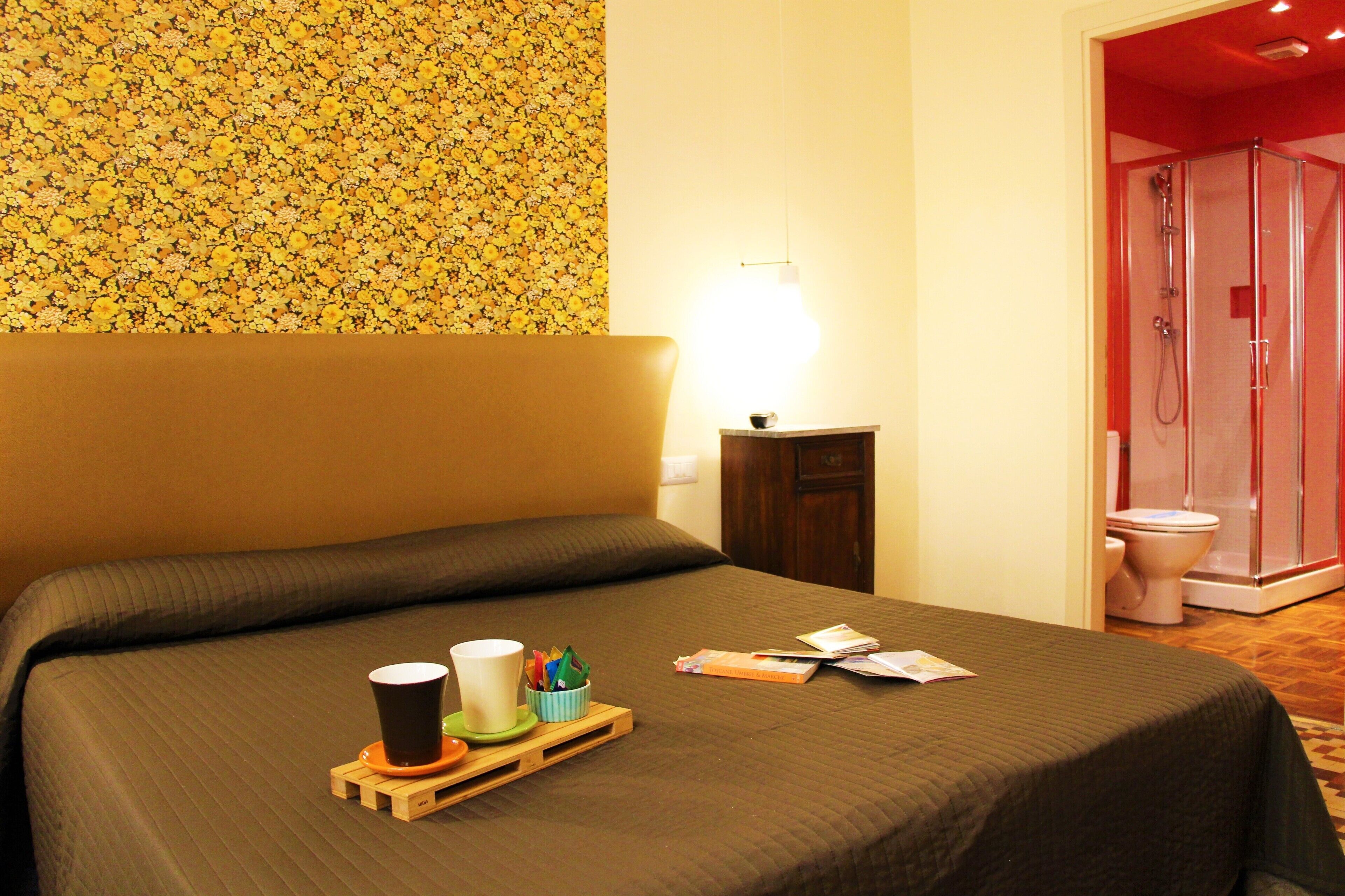 Quarto de hotel com uma cama, mesa de cabeceira iluminada por uma luminária e área de chuveiro visível através de uma porta aberta.