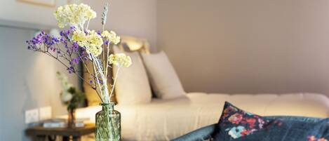 Um quarto com uma cama, uma cadeira com almofada florida, uma mesinha com um vaso de flores e duas fotos emolduradas na parede.