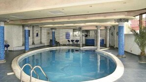 Piscina interior com chão revestido de azulejos, colunas e área de recepção com cadeiras.