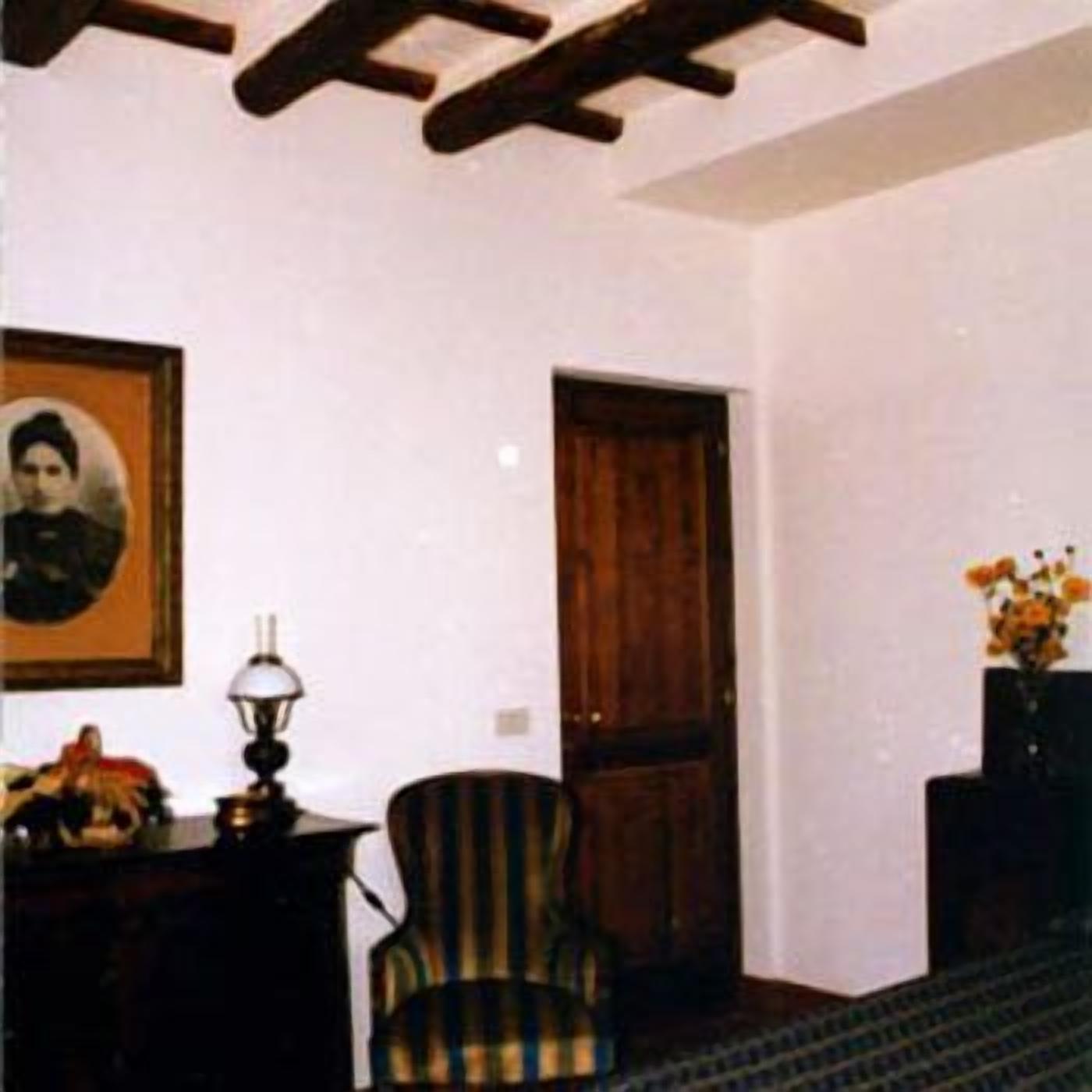 Quarto com teto de madeira, retrato emoldurado na parede, porta de madeira, poltrona listrada, mesa com abajur e um vaso com flores.
