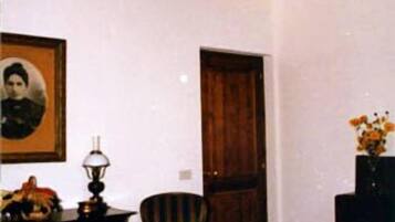 Quarto com teto de madeira, retrato emoldurado na parede, porta de madeira, poltrona listrada, mesa com abajur e um vaso com flores.