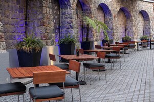 Um café ao ar livre com mesas e cadeiras de madeira, plantas em vasos e uma parede de pedra com arcos.