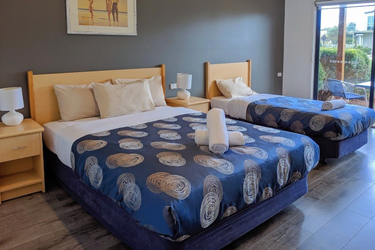 Quarto de hotel com duas camas, uma mesa de cabeceira com abajur, um quadro na parede e vista pela janela grande.