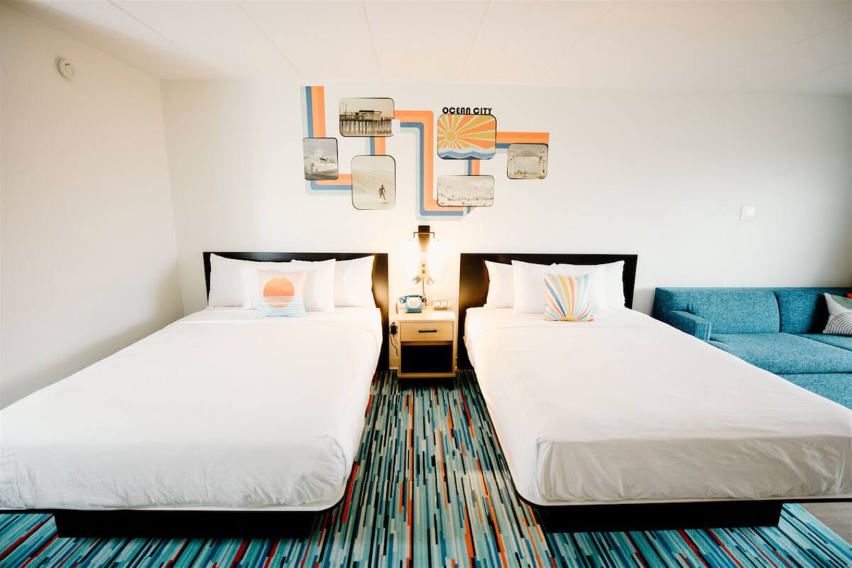 Quarto de hotel com duas camas, uma mesa de cabeceira, uma luminária e uma obra de arte na parede com a inscrição 'OCEAN CITY'.