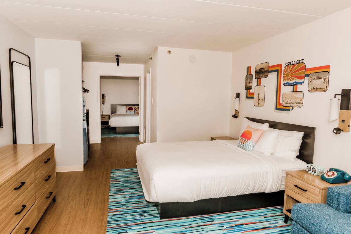 Quarto de hotel moderno com uma cama grande, uma cômoda de madeira, uma mesa de cabeceira com telefone e um tapete listrado e colorido.