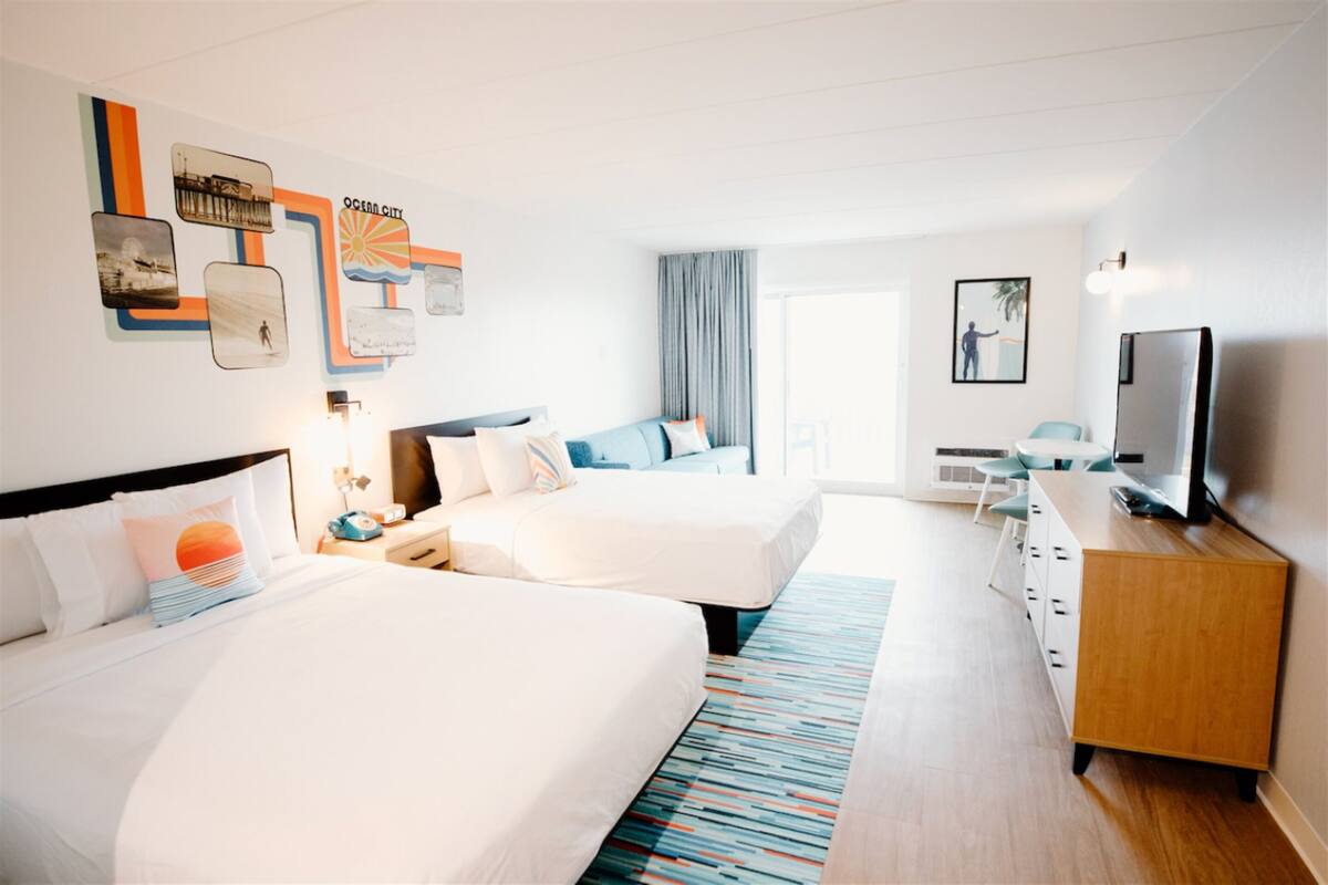 Quarto de hotel com duas camas, uma escrivaninha, televisão e obras de arte nas paredes.
