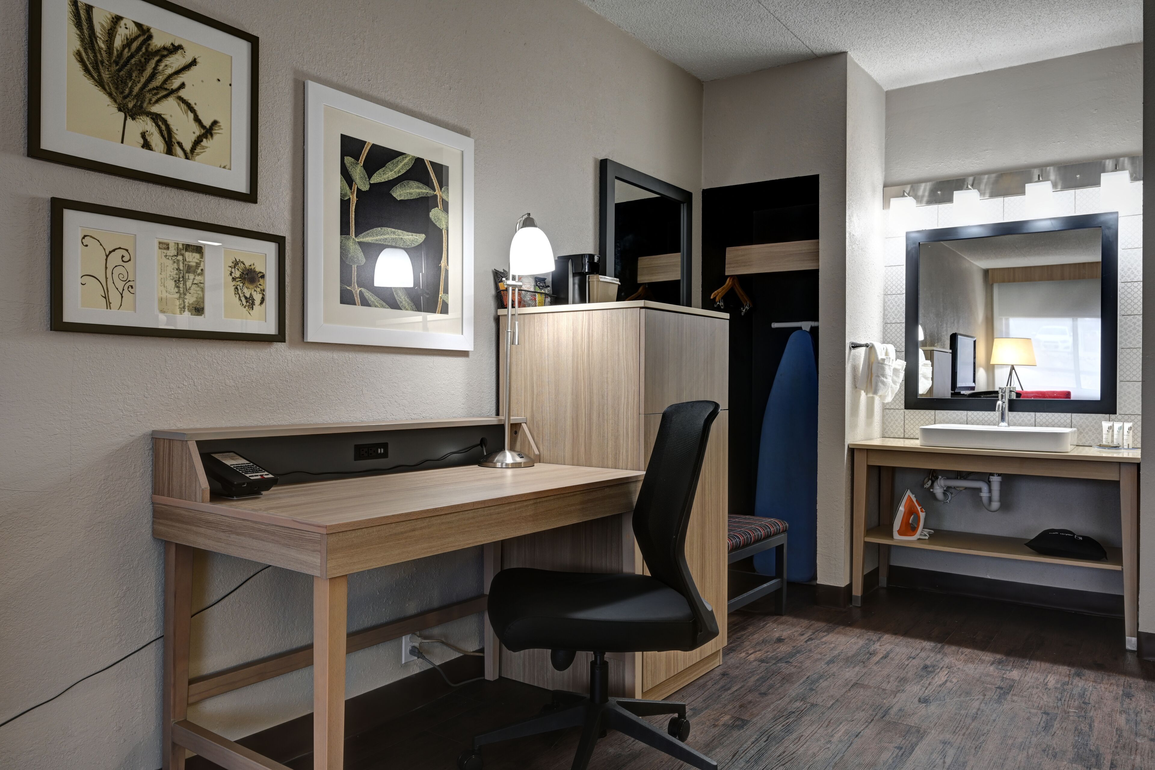 Quarto de hotel com uma escrivaninha de madeira, uma cadeira de escritório preta, um espelho, uma pia e obras de arte emolduradas na parede.