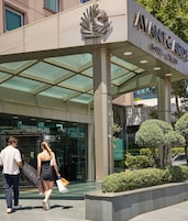 Entrada de hotel com fachada de vidro moderna, um logotipo em destaque e área ajardinada bem cuidada com arbustos.