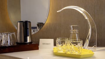 Mini-bar de hotel com cafeteira, jarra de vidro e outra jarra de vidro sobre um tabuleiro amarelo.