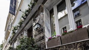 Fachada de hotel com placa 'HOTEL MUGUET', grandes janelas e caixas de flores.