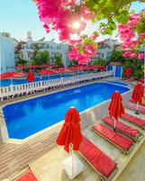 Uma piscina com guarda-sóis vermelhos e espreguiçadeiras, rodeada por edifícios e flores cor-de-rosa.