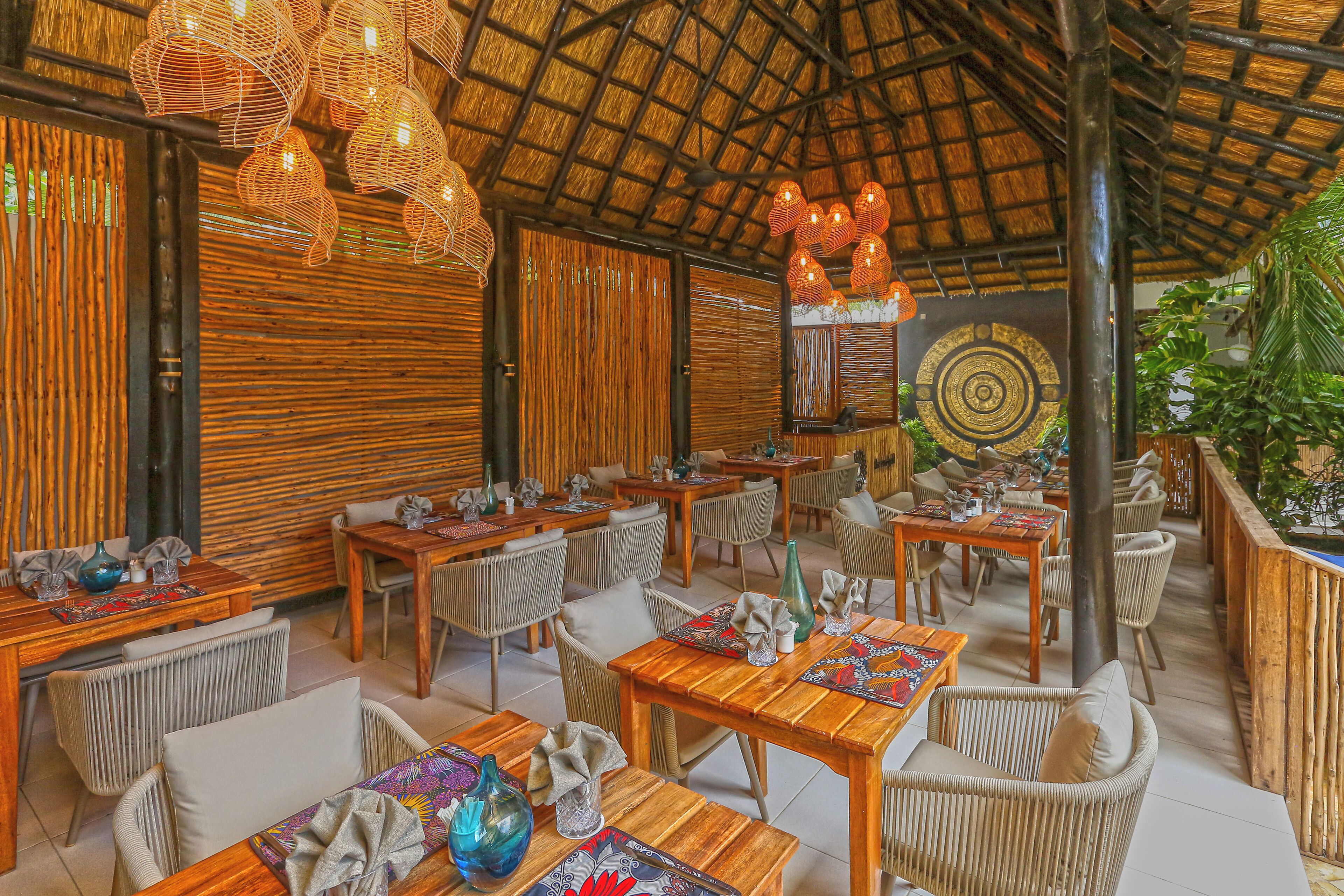 Um restaurante interno com mesas e cadeiras de madeira, teto de palha e luminárias de vime suspensas.