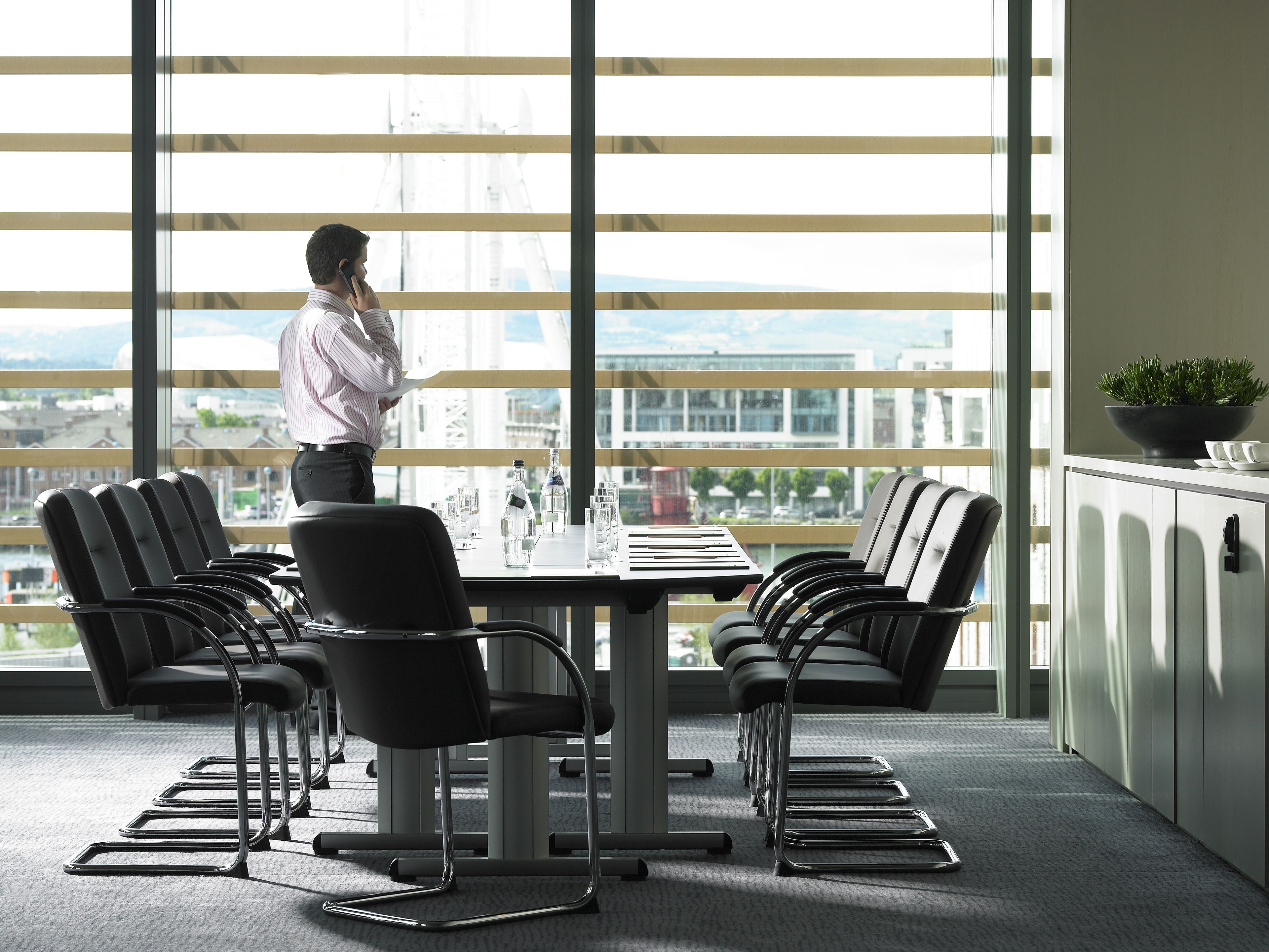 Uma sala de conferências com uma mesa grande e cadeiras, janelas amplas com persianas e vista para edifícios.