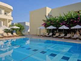 Piscina com espreguiçadeiras e guarda-sóis, cercada por edifícios e plantas floridas.