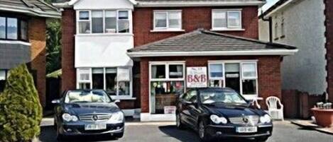 Um edifício de dois andares com uma placa 'B&B' indicando ser um alojamento local. Dois carros estão estacionados em frente.