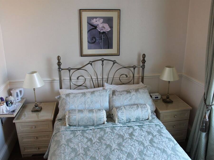 Uma cama bem arrumada com colcha estampada, duas mesas de cabeceira com luminárias, um quadro na parede e uma janela com cortinas.
