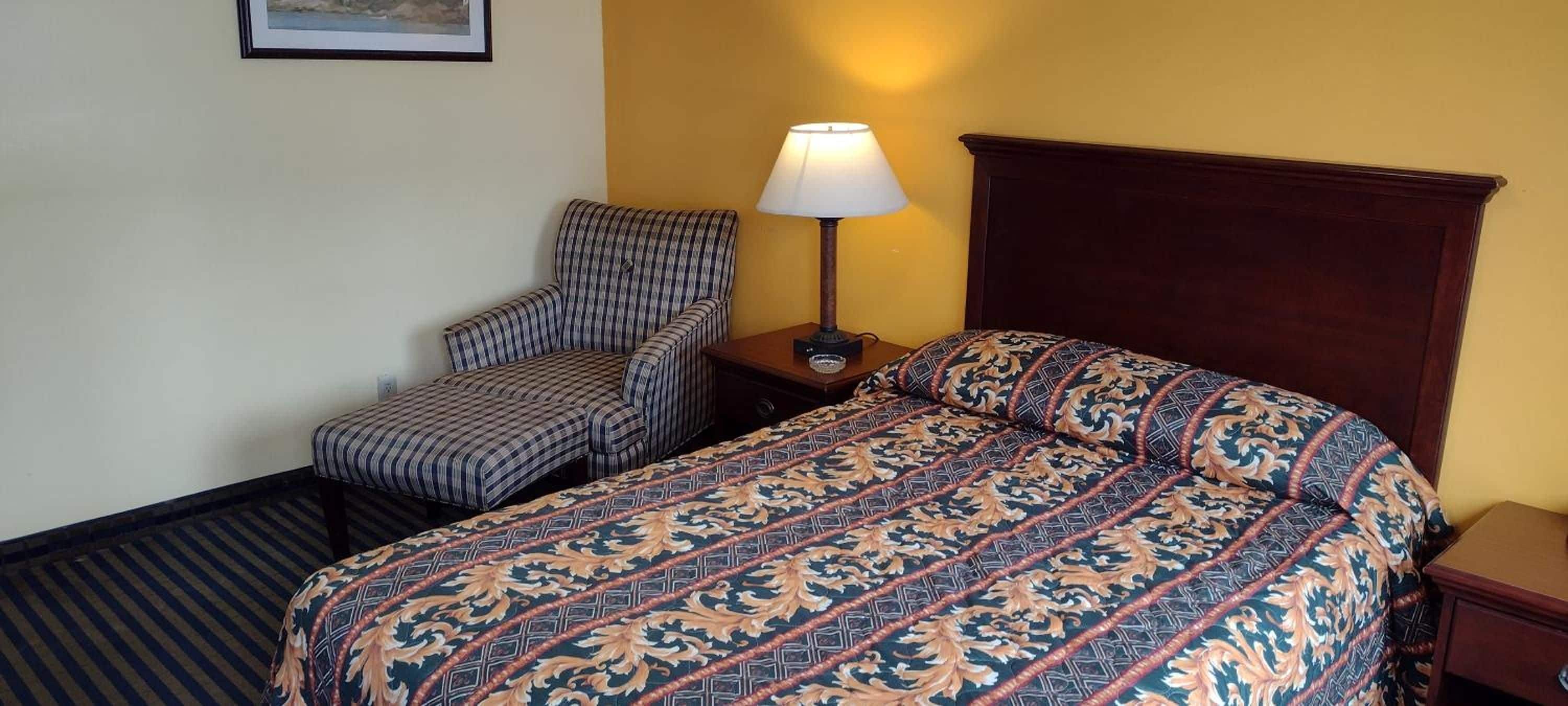 Quarto de hotel com cama, uma cadeira, mesa de cabeceira e abajur.