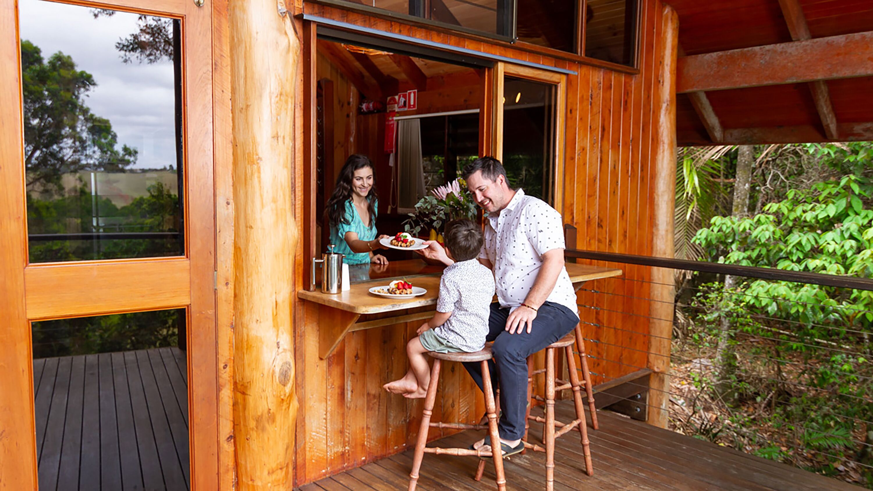 Uma cabana de madeira com varanda, área de jantar com mesa e cadeiras, e janela com vista para vegetação.