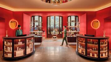 Um hall com esquema de cores vermelho, duas vitrines de vidro, grandes janelas e um lustre.