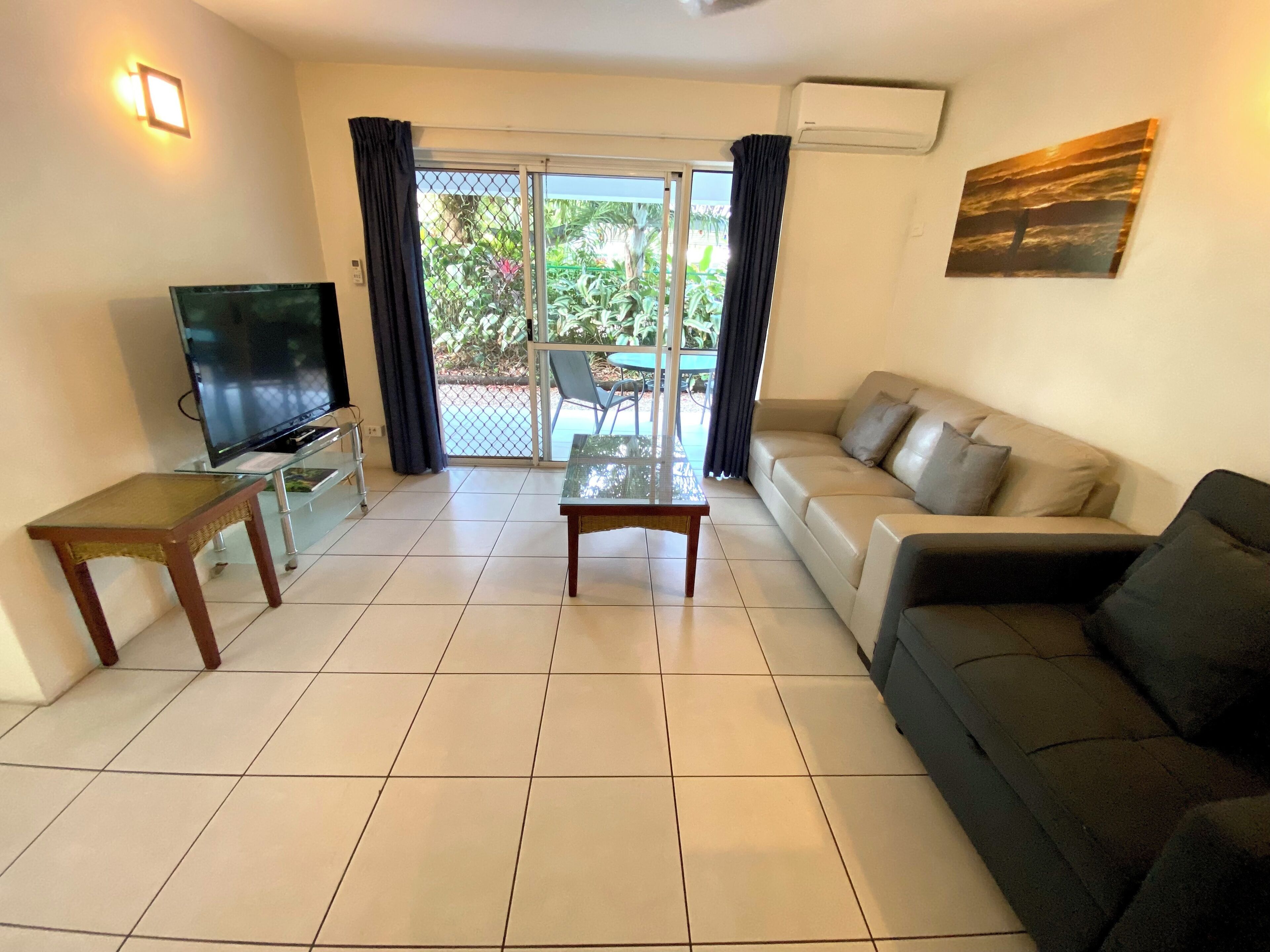 Uma sala de estar com televisão, sofá, mesa de centro e uma porta de vidro deslizante que dá para uma área externa com cadeiras.