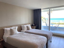 Quarto de hotel com duas camas, uma janela grande com vista para o mar e uma varanda com cadeiras.