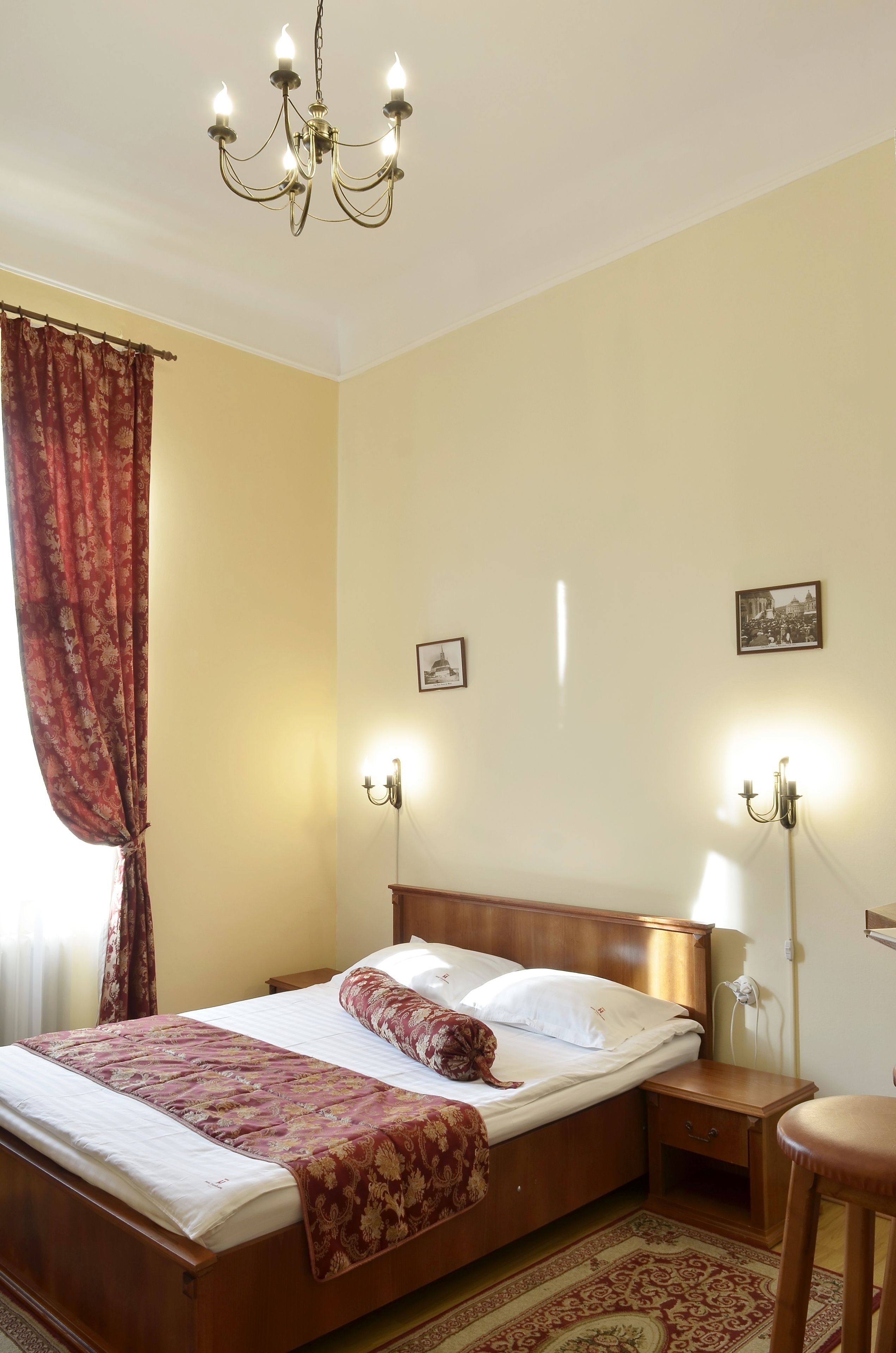 Uma cama bem arrumada com colcha estampada e almofadas, uma mesa de cabeceira de madeira com abajur, e um candelabro acima.