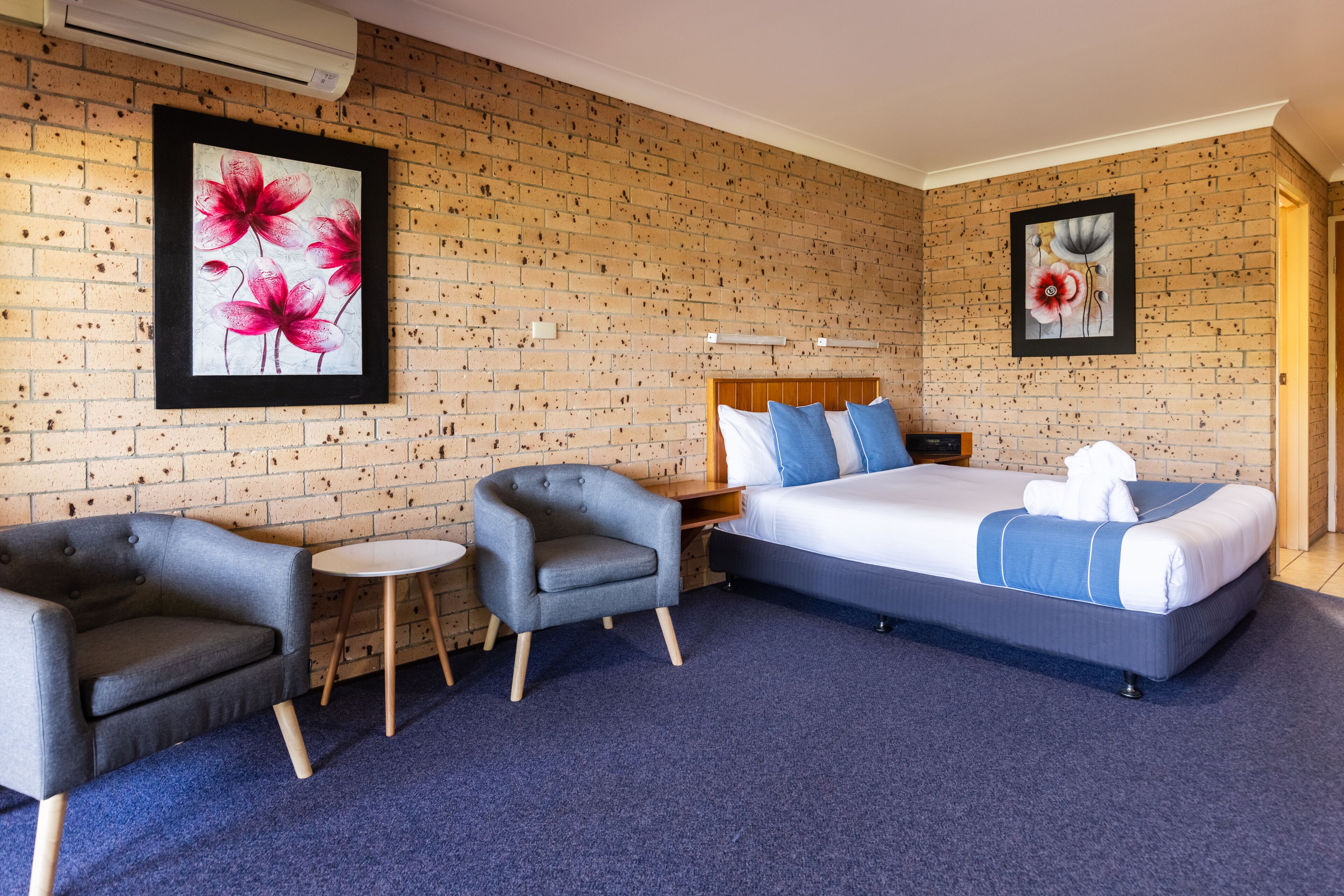 Quarto de hotel com parede de tijolos, cama com roupa de cama azul, poltrona cinza, mesa redonda pequena e duas obras de arte florais emolduradas.